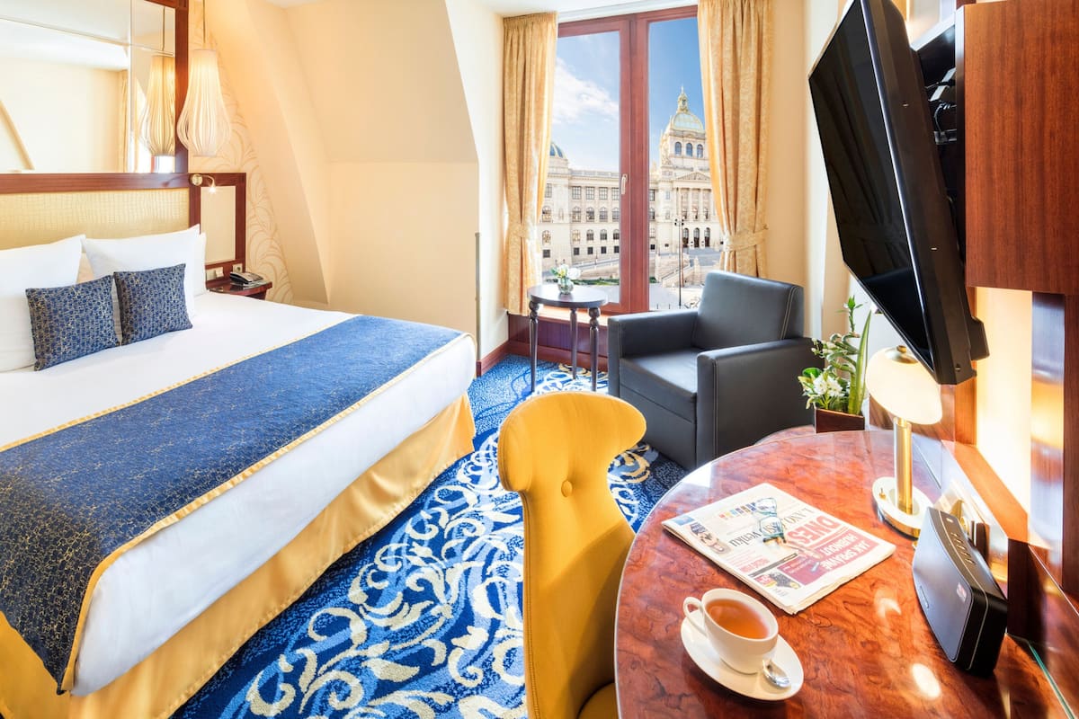 Quarto de hotel com uma cama grande, uma escrivaninha com chá, uma cadeira e uma televisão.