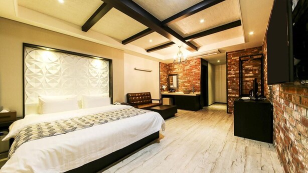 Quarto de hotel moderno com uma cama grande, área de estar com um sofá, uma pequena mesa de escritório e uma parede de tijolos decorativa.