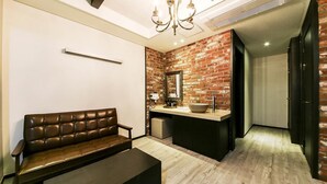 Um interior moderno com sofá de couro, um banquinho preto, parede de tijolo, piso de madeira e um banheiro pequeno com pia e espelho.