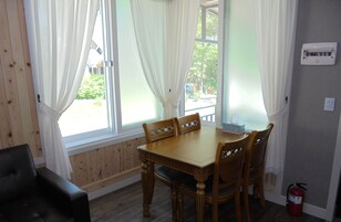 Uma área de refeições com mesa e cadeiras de madeira, janelas amplas com cortinas brancas, um extintor de incêndio e um termostato na parede.