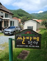 Um imóvel residencial com um carro estacionado em frente, uma casa pequena e uma placa com informações de contato.