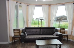 Quarto com sofá de couro escuro, mesa de centro de madeira e duas cadeiras de madeira. Janelas grandes com cortinas brancas e vista para o verde lá fora.