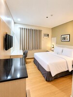 Quarto de hotel com cama, escrivaninha, cadeira, televisão de tela plana, janela com cortinas, aparelho de ar condicionado e quadro na parede.