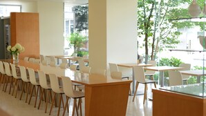 Uma área de jantar moderna com mesas e cadeiras longas, amplas janelas com vista para o exterior e um balcão central de madeira.