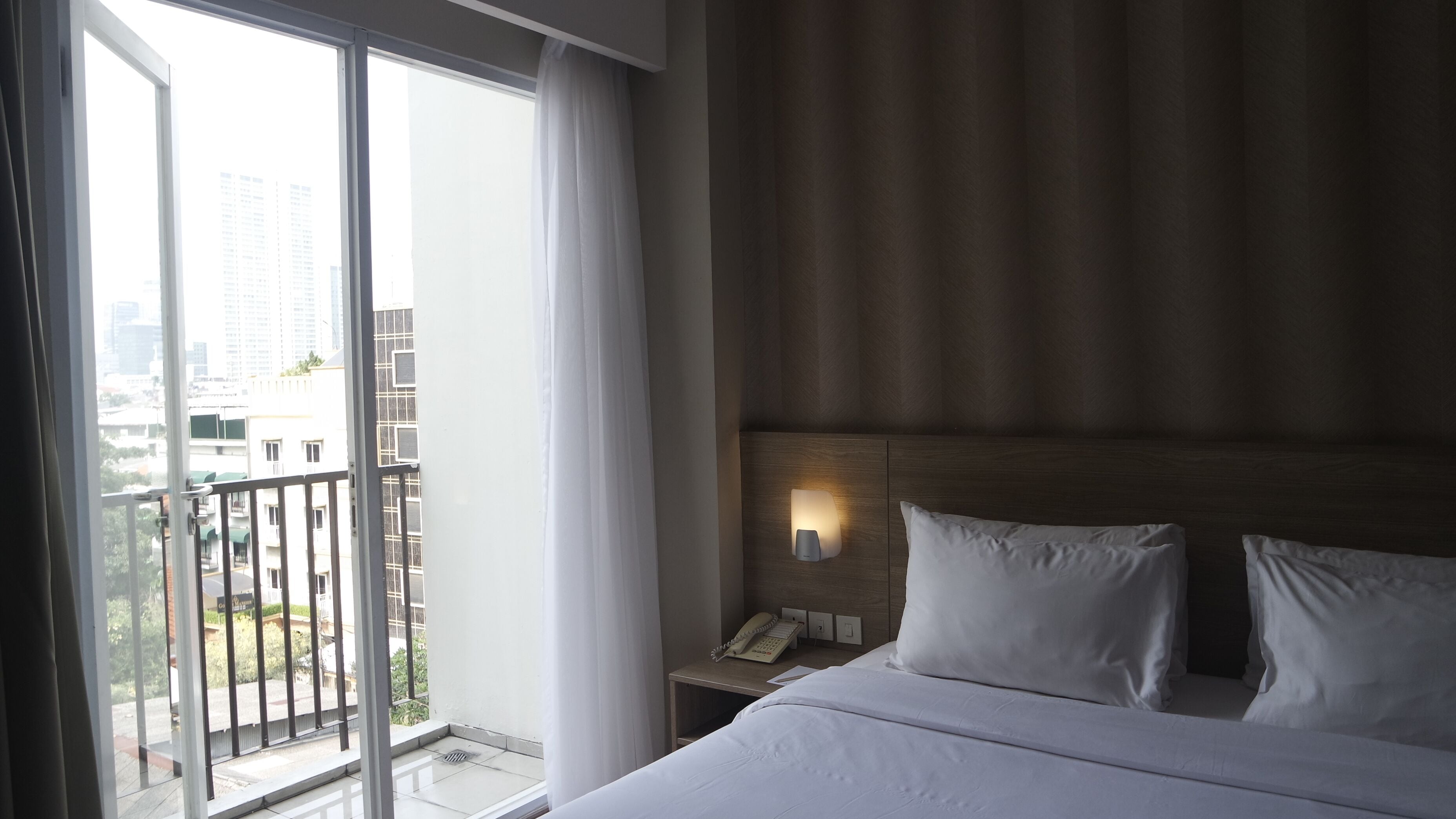 Quarto de hotel com uma janela grande, cama com roupa de cama branca, mesa de cabeceira com abajur e telefone.