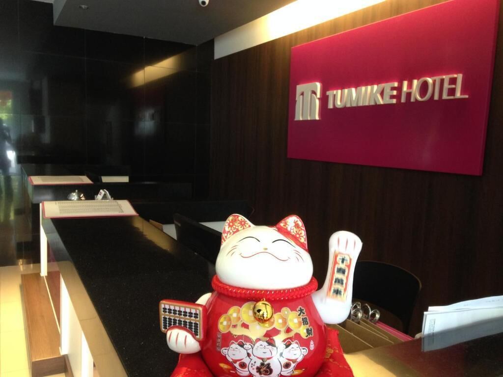 Uma mesa de recepção com uma placa vermelha do Hotel Tumike, uma estátua decorativa de gato e um balcão com uma máquina registradora.