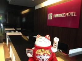 Uma mesa de recepção com uma placa vermelha do Hotel Tumike, uma estátua decorativa de gato e um balcão com uma máquina registradora.