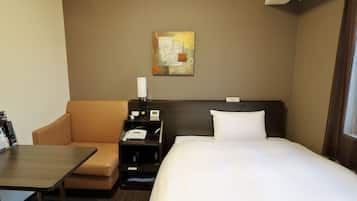 Quarto de hotel com cama, escrivaninha, cadeira, mesa de cabeceira com telefone e luminária, e um quadro na parede.