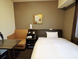 Quarto de hotel com cama, escrivaninha, cadeira, mesa de cabeceira com telefone e luminária, e um quadro na parede.
