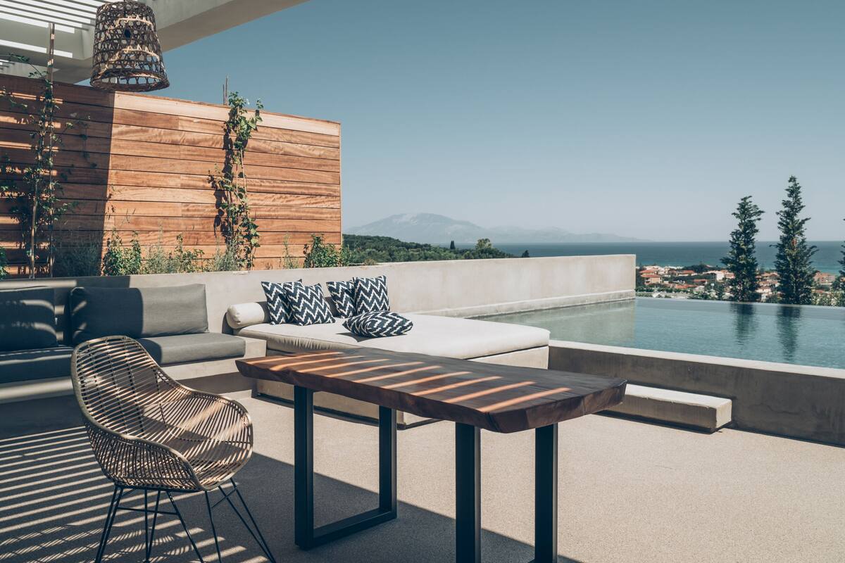 Uma área de estar moderna ao ar livre, com mesa de madeira, cadeira de vime e sofá com almofadas estampadas.