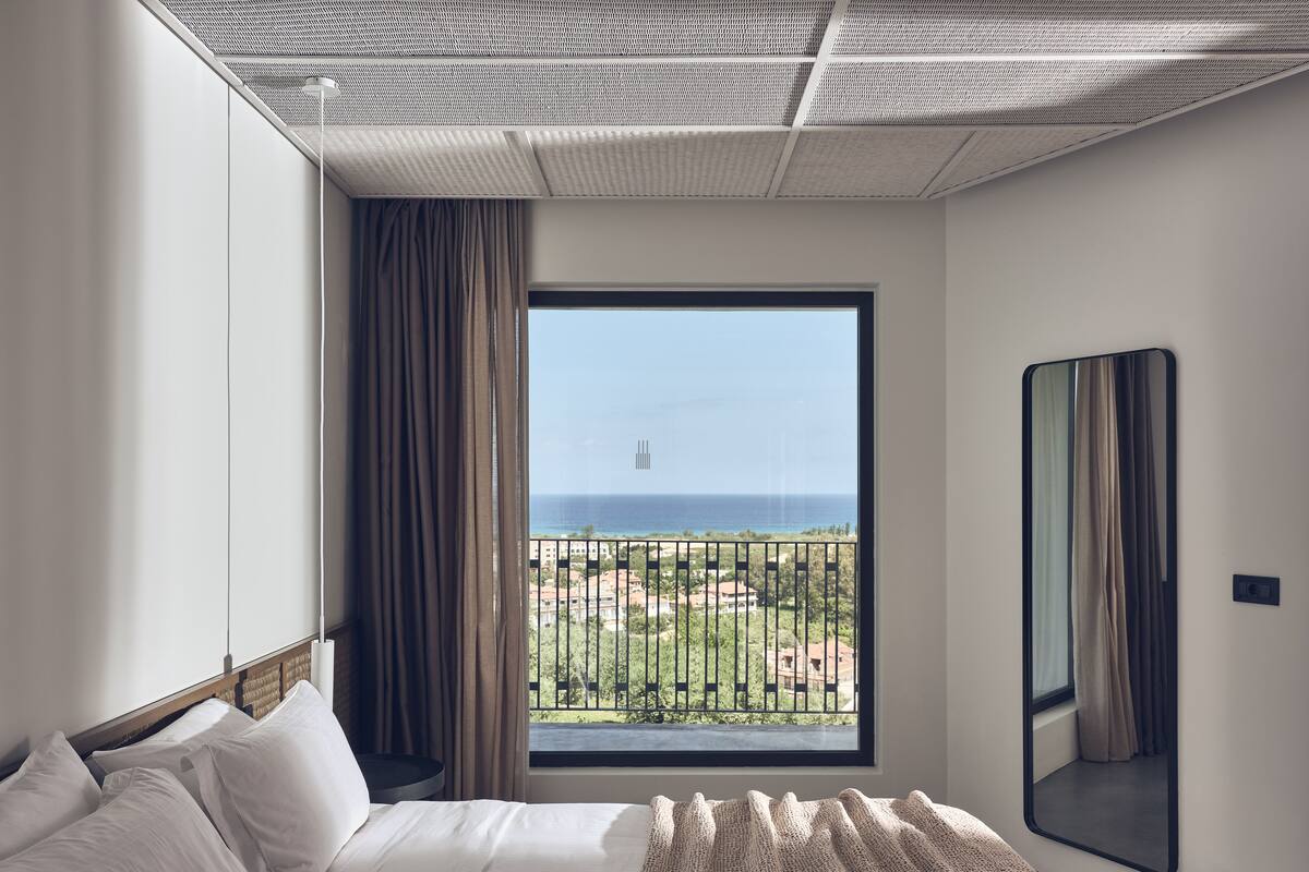 Quarto de hotel com uma janela grande, uma cama, uma mesa de cabeceira e um espelho.