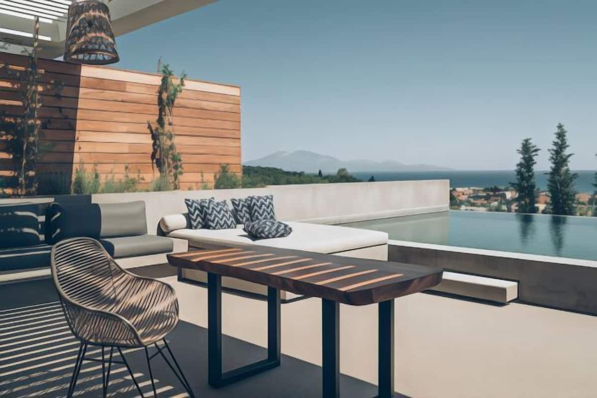 Uma área de estar moderna ao ar livre com piscina, parede de madeira e vista para o mar e montanhas.