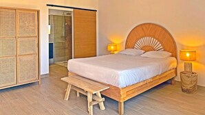Quarto com cama de madeira, duas luminárias de cabeceira, um banquinho, guarda-roupa de vime e porta de chuveiro de vidro.