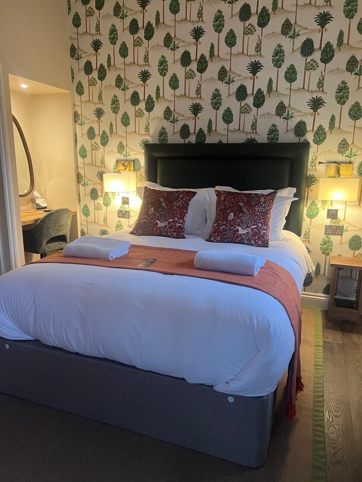 Uma cama bem arrumada com lençóis brancos e duas almofadas decorativas, uma cabeceira escura e uma parede com padrão de árvore.