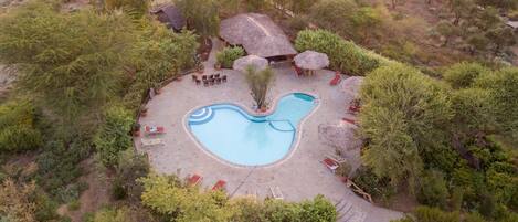 Vista aérea de um resort com uma piscina central, rodeada por espreguiçadeiras e construções com telhados de palha.