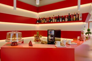 Uma área de bar vermelha e branca, com diversos tipos de bebidas, uma máquina de café e uma exposição de frutas.