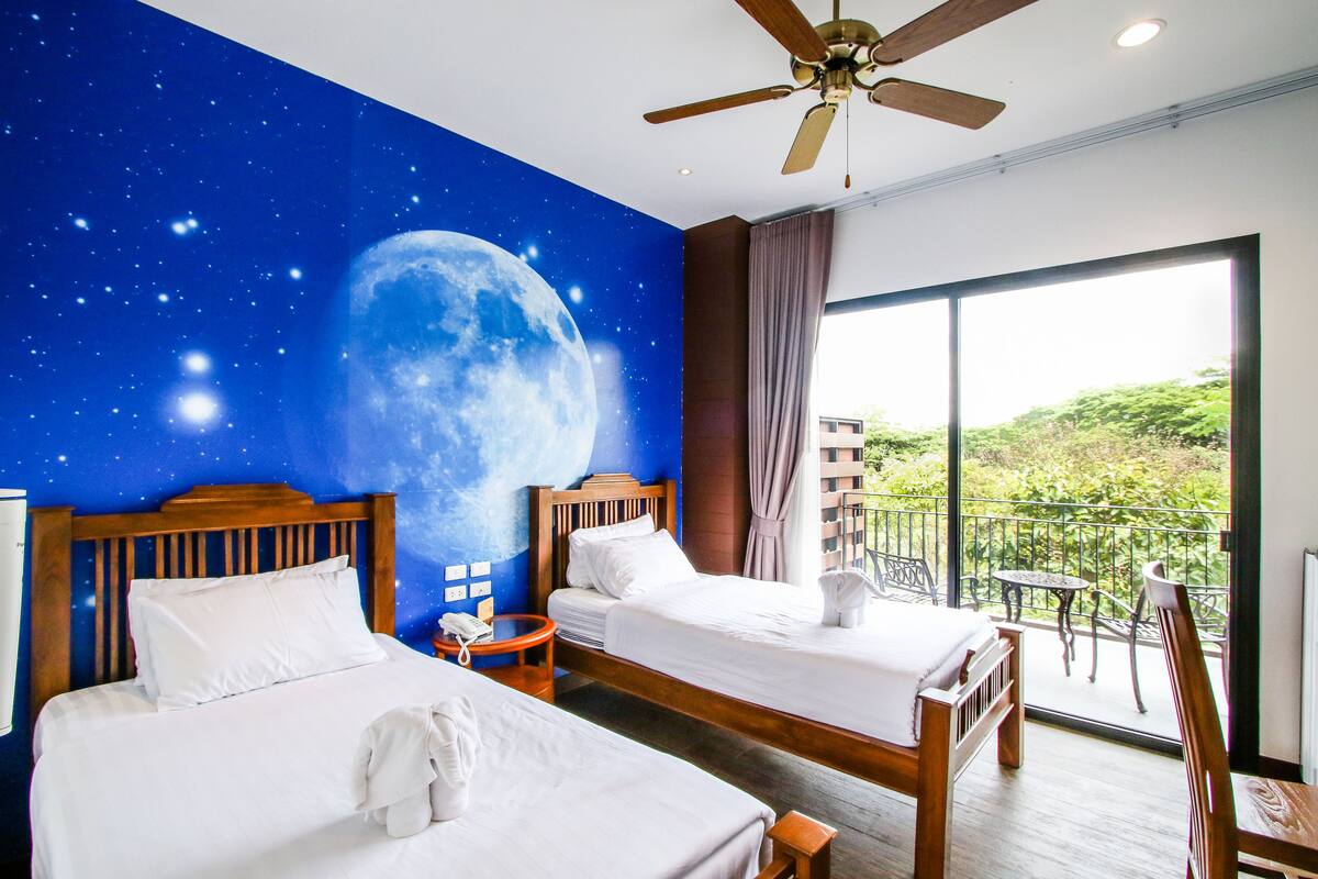 Quarto de hotel com duas camas, um ventilador de teto, uma janela grande com varanda e um mural de um planeta na parede.