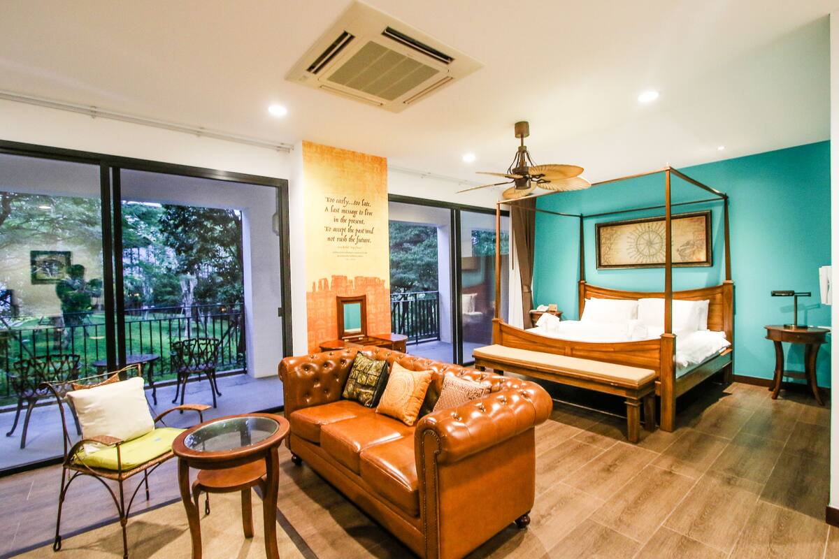 Quarto de hotel com uma cama grande, um sofá de couro, um banco de madeira e uma varanda com mobiliário exterior.