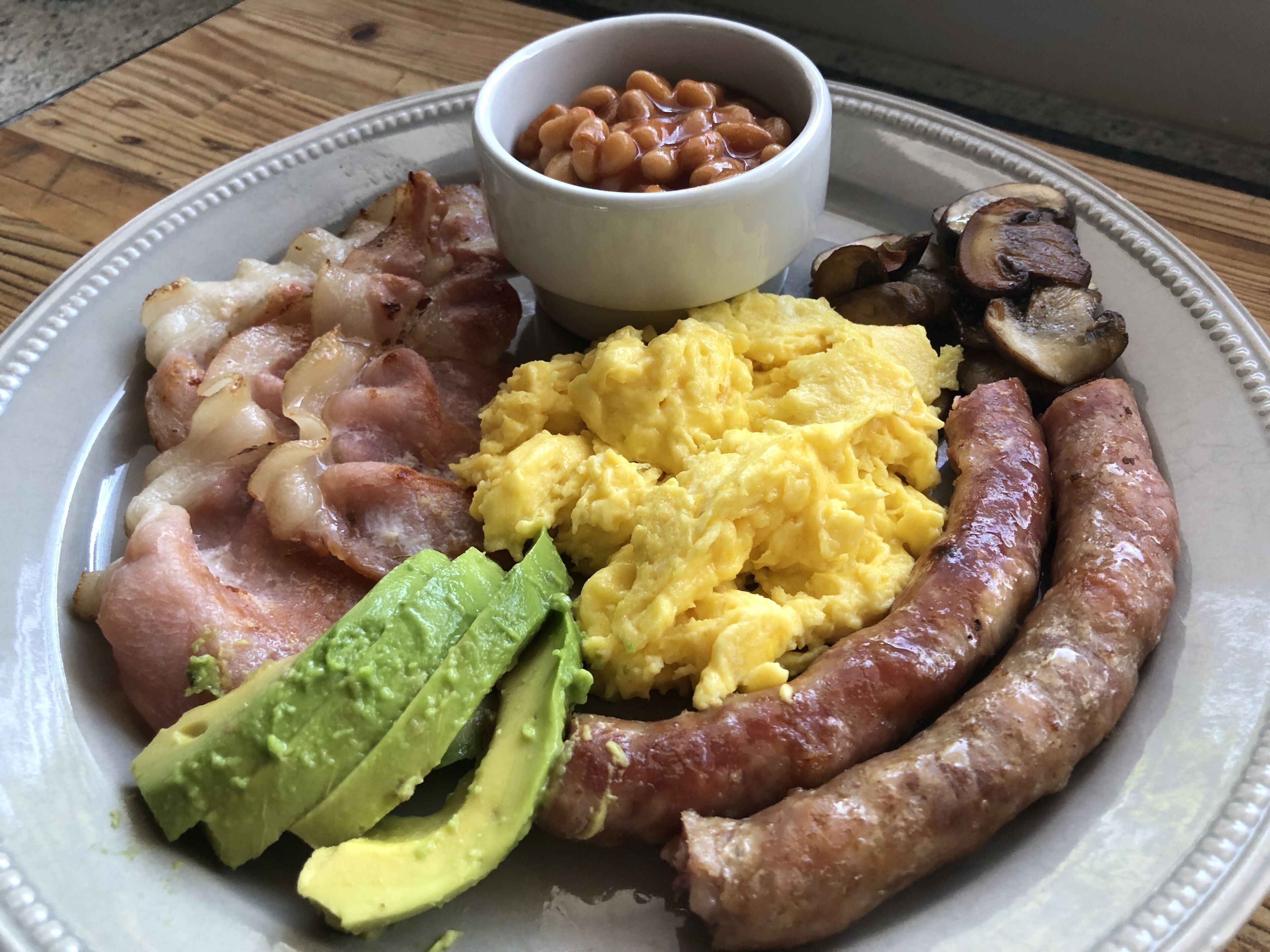 Um prato com bacon, salsichas, ovos mexidos, fatias de abacate, cogumelos e feijão cozido.