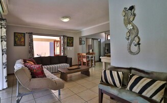 Sala de estar com sofá, poltrona e mesa de centro. Há um espaço de refeições com cadeiras e mesa. É visível uma peça decorativa na parede.