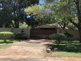 Uma entrada protegida por portão, com uma longa entrada de automóveis que leva a um edifício, cercado por árvores e gramados bem cuidados.