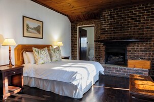 Quarto com cama grande, cabeceira de madeira, duas mesas de cabeceira com luminárias, lareira de tijolos e um quadro na parede.