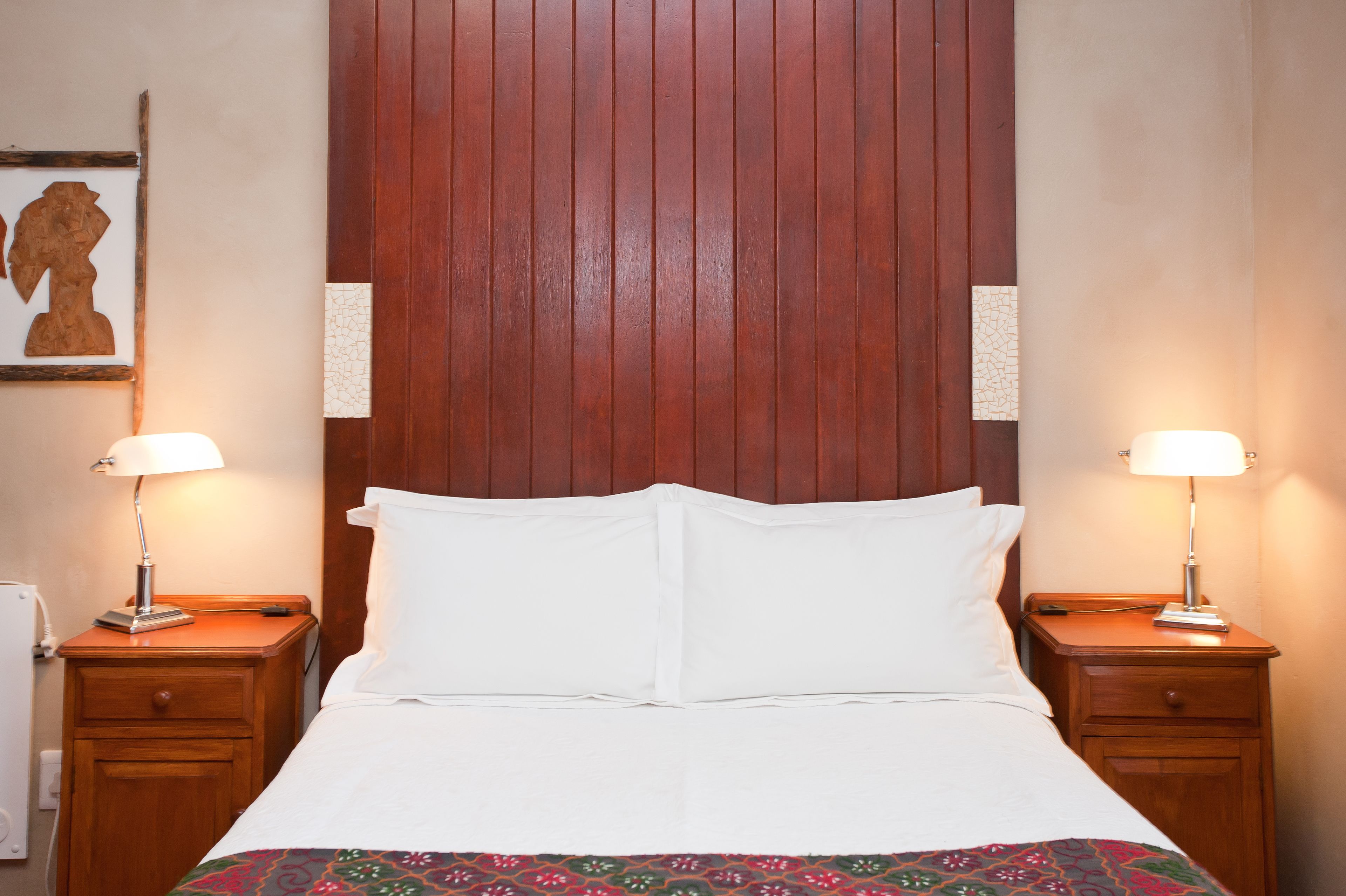 Quarto de hotel com cabeceira de madeira, duas mesas de cabeceira com abajures, cama com roupa de cama branca e manta estampada ao pé da cama.