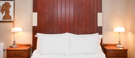 Quarto de hotel com cabeceira de madeira, duas mesas de cabeceira com abajures, cama com roupa de cama branca e manta estampada ao pé da cama.