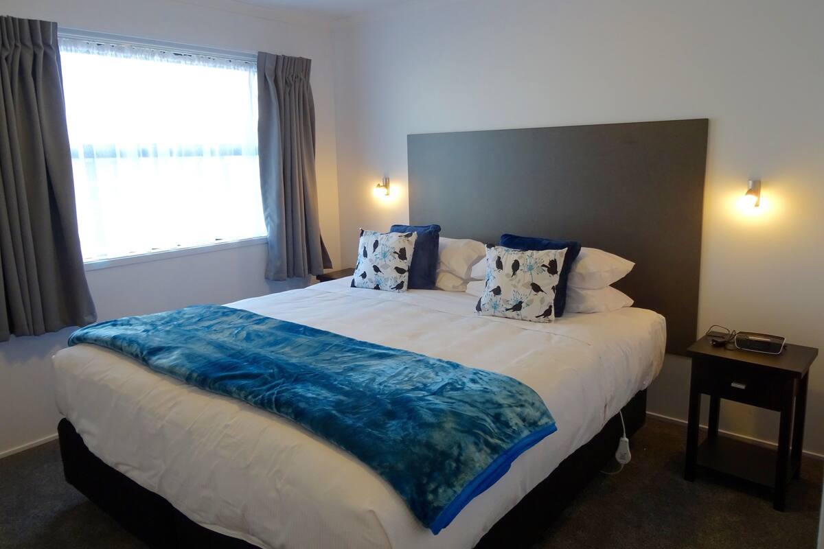 Quarto de hotel com uma cama grande, uma mesa de cabeceira com abajur e uma janela com cortinas.