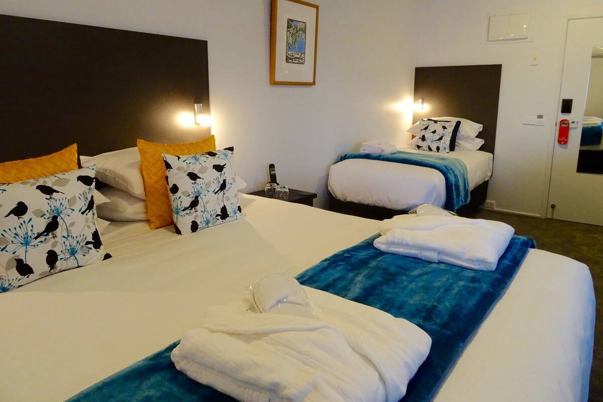 Quarto de hotel com duas camas, cada uma com lençóis brancos e cobertores azuis. Há almofadas decorativas, um quadro na parede e uma mesa de cabeceira com um telefone e óculos.
