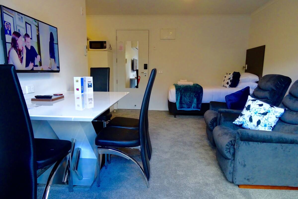 Quarto de hotel com uma mesa de jantar, cadeiras, um sofá, uma cama com cobertor e uma televisão exibindo um programa.