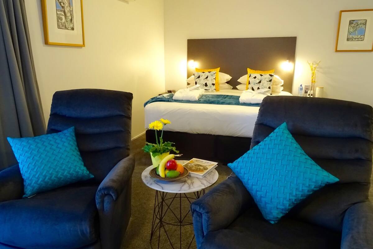 Quarto de hotel com cama, duas poltronas, uma mesinha redonda com frutas e um livro, e uma mesa de cabeceira com abajur.