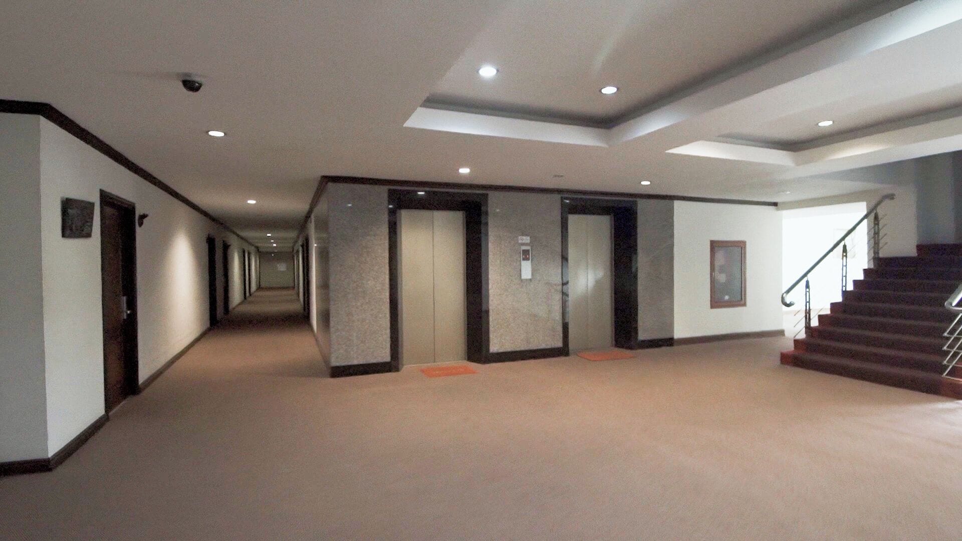 Corredor de hotel com piso carpeteado, escada e várias portas de acesso aos quartos.