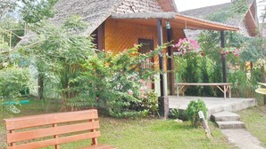 Um edifício rústico com telhado de colmo, rodeado por vegetação exuberante e um banco de madeira em primeiro plano.