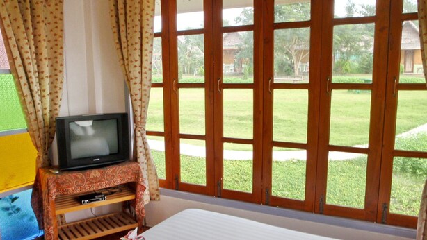 Quarto com cama, televisão sobre suporte de madeira e amplas janelas com vista para um gramado e árvores.