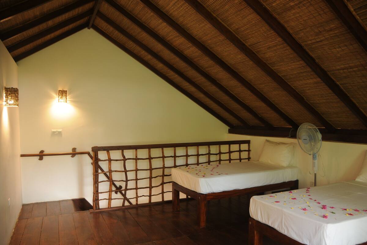 Quarto com duas camas, piso de madeira, teto inclinado e um ventilador.