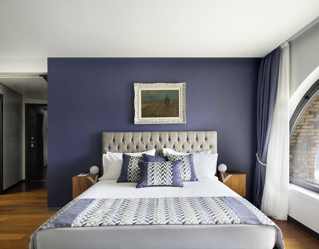 Quarto com cabeceira estofada, cama grande com roupa de cama estampada, quadro na parede e vista da cidade pela janela.