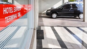 Entrada moderna de hotel com fachada de vidro, uma placa vermelha indicando terminal express do hotel e um carro preto estacionado no interior.