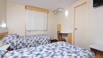 Quarto de hotel com duas camas, uma escrivaninha, uma cadeira, ar condicionado e uma televisão na parede.