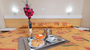Uma bandeja de café da manhã bem organizada com doces, uma fruta, uma xícara de café e um vaso decorativo sobre uma cama com colcha estampada e cabeceira de madeira.