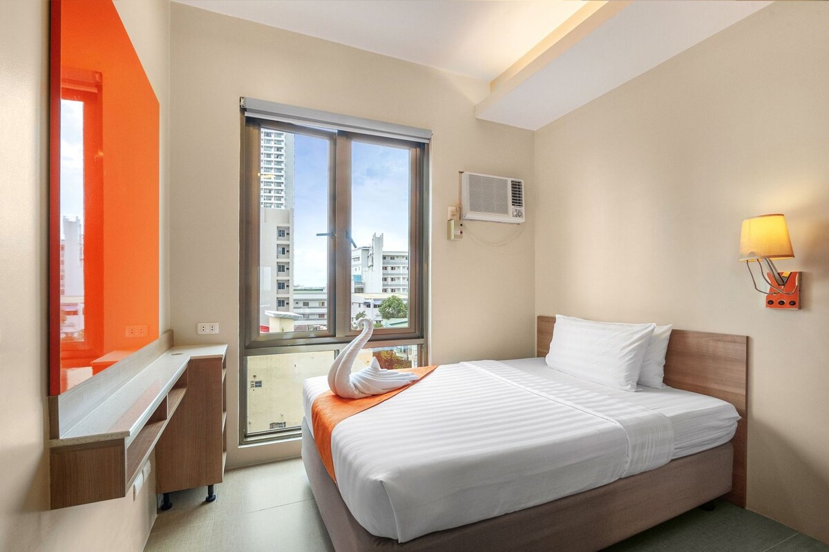 Quarto de hotel com cama, escrivaninha, cadeira, janela e vista para edifícios, e unidade de ar condicionado.