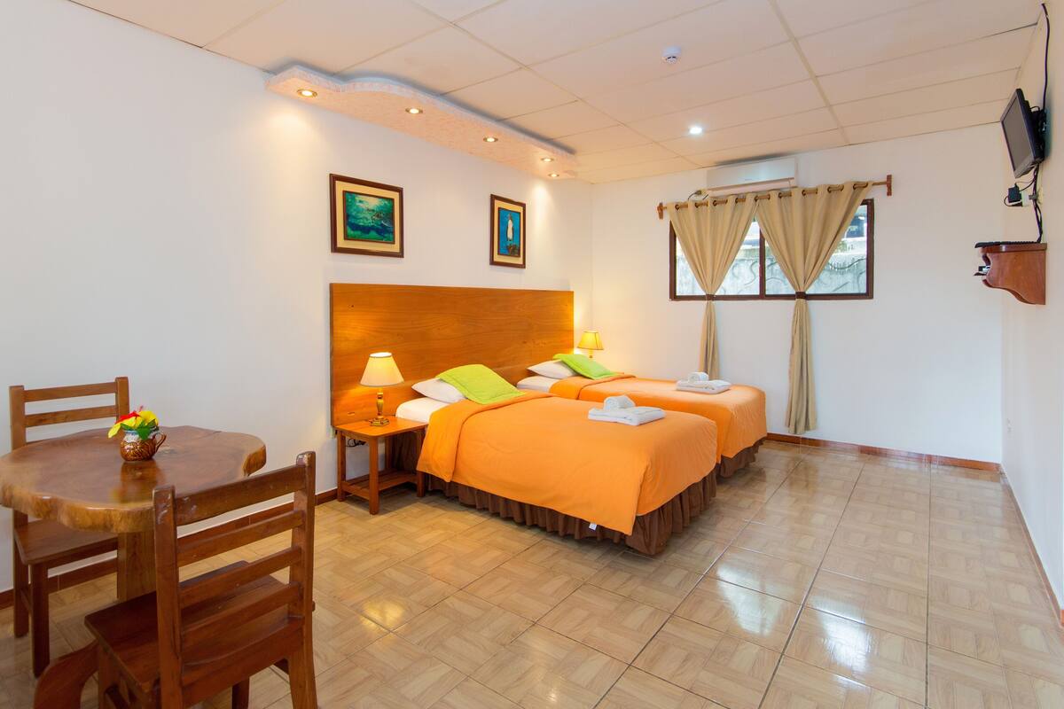 Quarto de hotel com cabeceira de madeira, duas camas com roupa de cama laranja, uma mesa de jantar de madeira com cadeiras e uma mesinha de cabeceira com abajur.