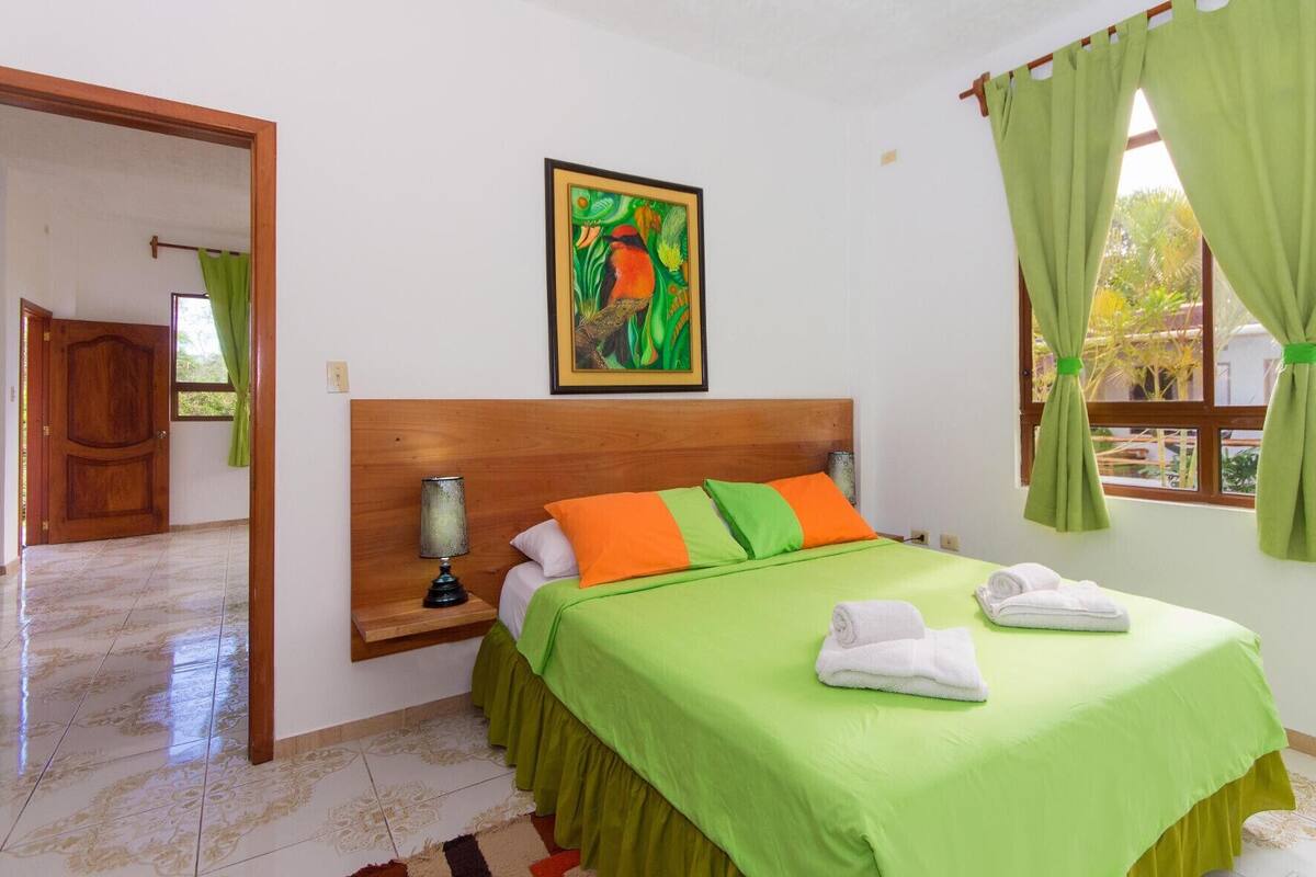 Quarto de hotel com cama, mesa de cabeceira, luminária, pintura e janela com cortinas.