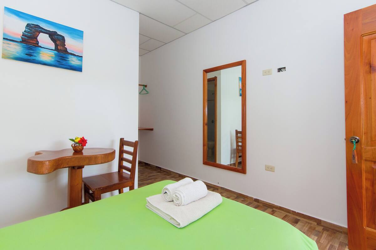 Quarto de hotel com cama, uma cadeira de madeira, uma mesa pequena com um vaso de flores, um quadro de uma formação rochosa e um espelho na parede.