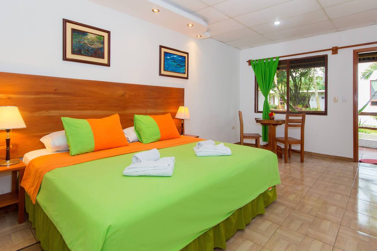 Quarto de hotel com cabeceira de madeira, duas pinturas emolduradas, colcha verde, almofadas laranja e verde, mesa de cabeceira com abajur, uma cadeira e uma janela com cortinas verdes.