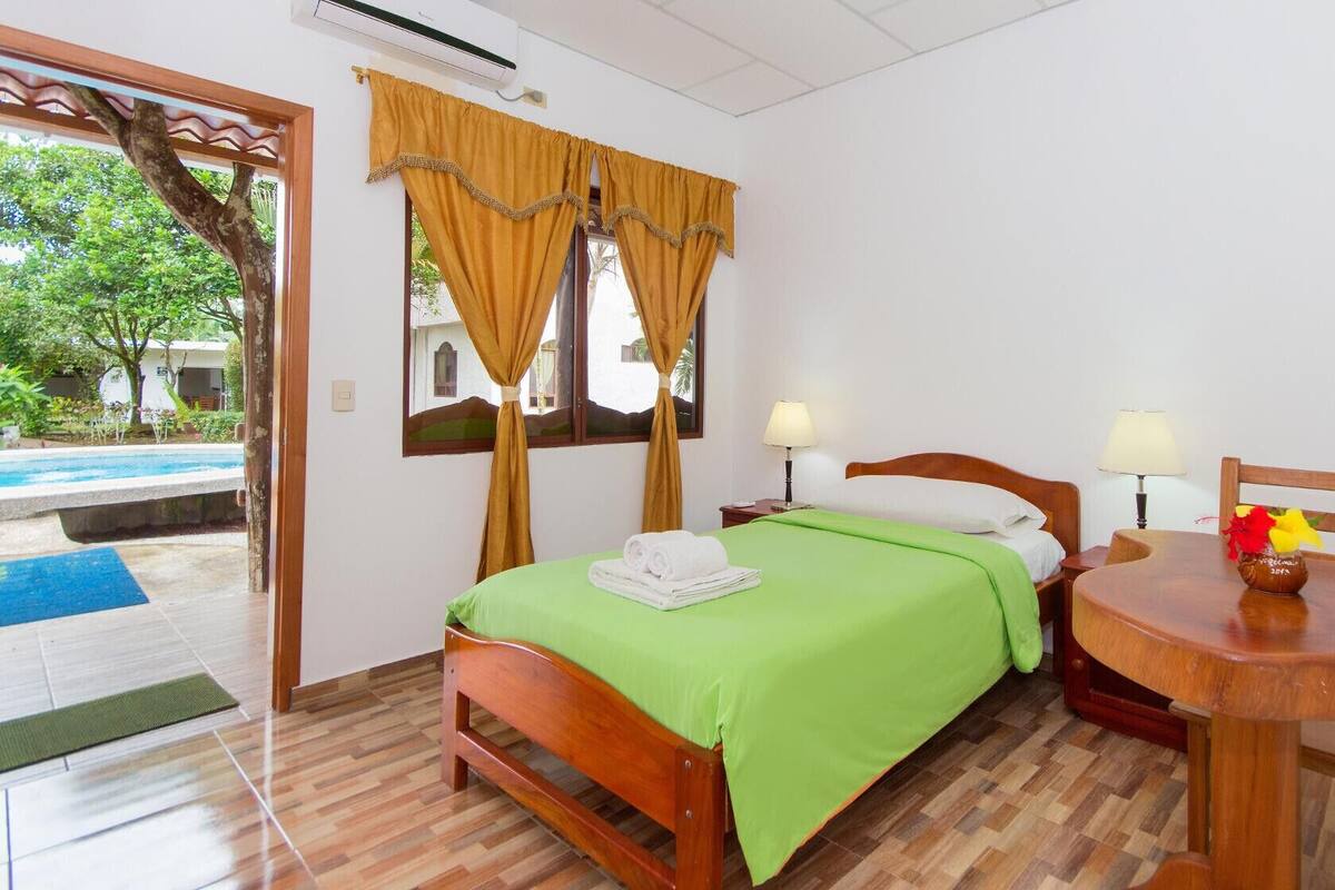 Quarto de hotel com uma cama, duas mesas de cabeceira com abajures, uma cadeira de madeira, uma pequena mesa com um vaso, e vista para uma piscina e vegetação lá fora.