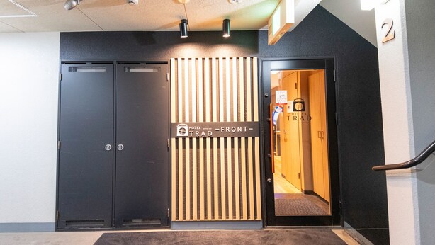 Entrada de hotel com design moderno, apresentando uma porta preta, um painel de madeira com o nome do hotel e piso coberto de carpete.
