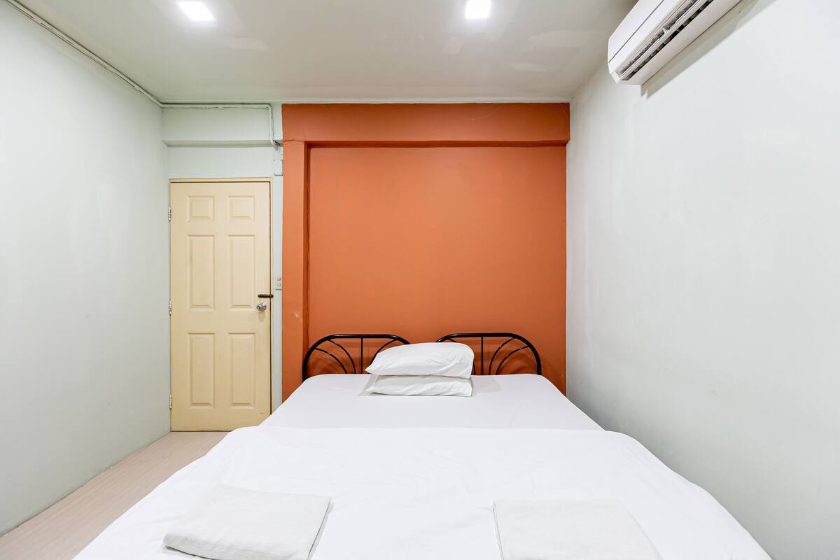 Um quarto pequeno, com cama de solteiro, uma cama simples, uma porta e um aparelho de ar condicionado na parede.