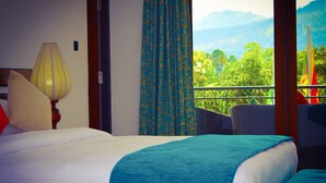 Quarto de hotel com cama, mesa de cabeceira, abajur e janela com vista para áreas verdes e montanhas.