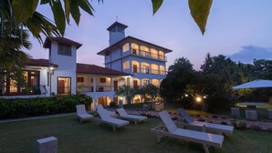 Um edifício de vários andares com varanda, rodeado por um gramado bem cuidado e área externa com mesas e cadeiras.